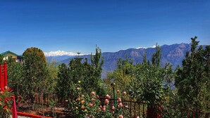 Um jardim com cadeiras vermelhas e um gazebo, rodeado por vegetação exuberante e vista para montanhas nevadas.