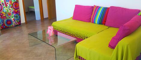 Quarto com sofá verde, almofadas cor-de-rosa e uma mesa de centro de vidro.