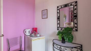 Um quarto com parede rosa, uma geladeira branca, uma cadeira, uma mesinha com uma planta e um espelho com moldura decorativa.