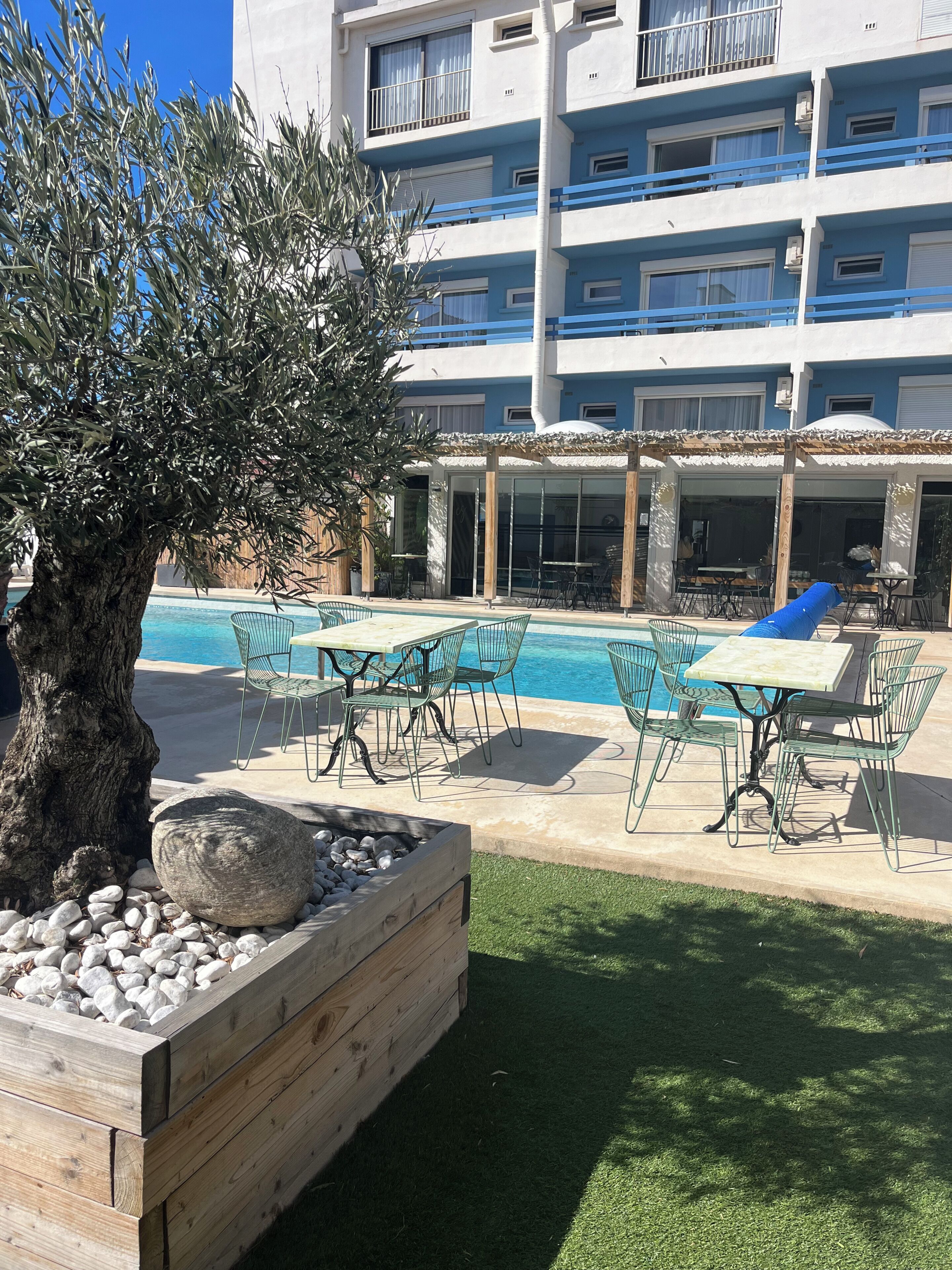 Uma área de piscina com mobiliário ao ar livre, uma grande árvore e um edifício de vários andares ao fundo.