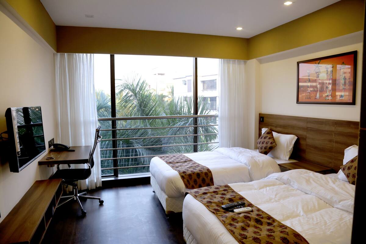 Quarto de hotel com duas camas, uma escrivaninha, uma cadeira, televisão e uma janela grande com vista para palmeiras.
