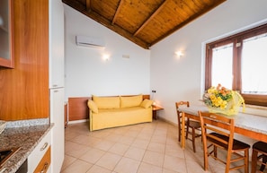 Cozinha compacta com teto de madeira, sofá amarelo, mesa de jantar com cadeiras e janela com flores no parapeito.