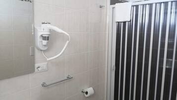 Casa de banho com secador de cabelo fixado na parede, um porta-toalhas, um rolo de papel higiénico e zona de duche com porta de vidro.