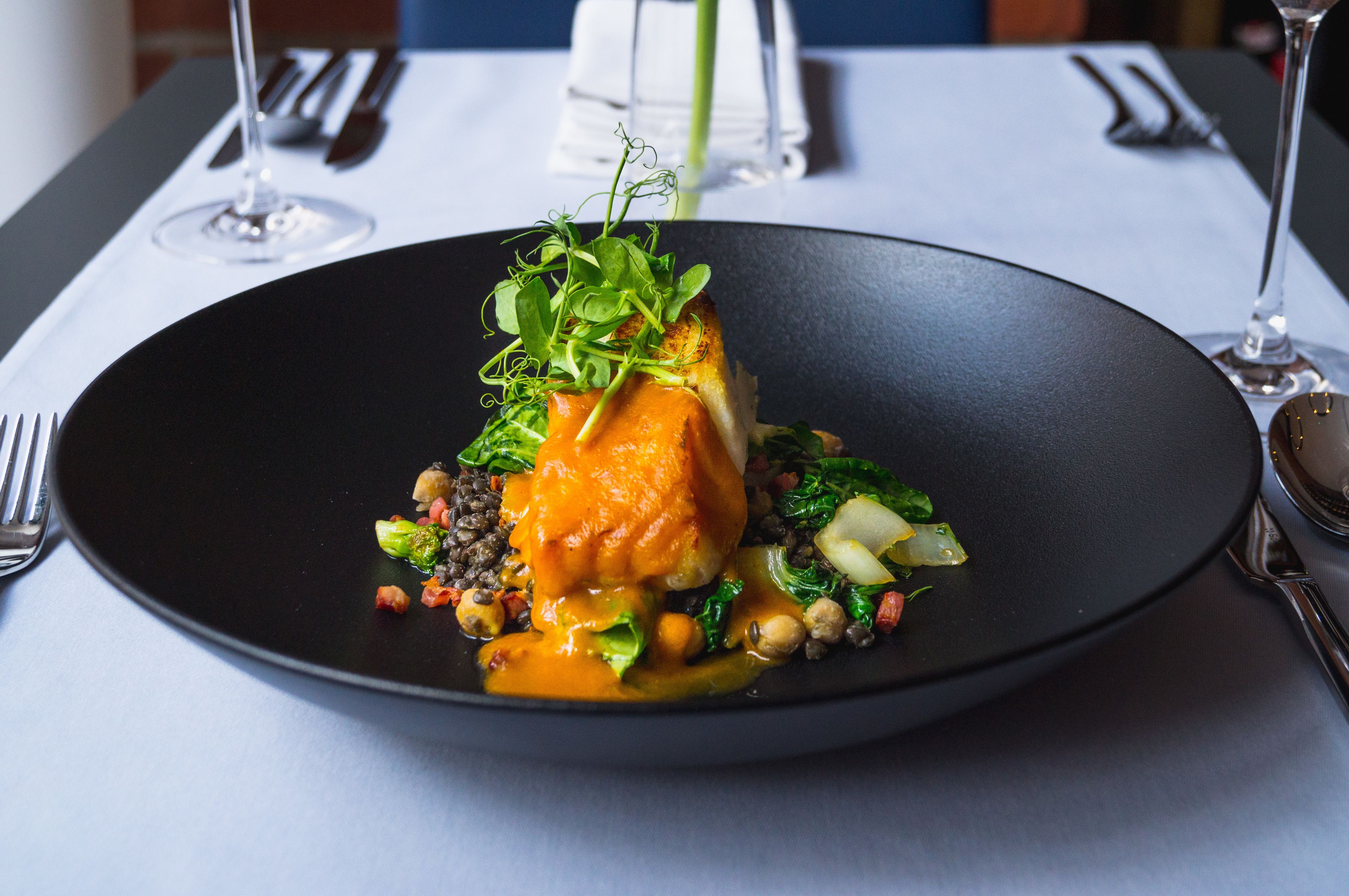 Um prato preto com um prato de peixe, legumes e vegetais sobre uma mesa com toalha branca.