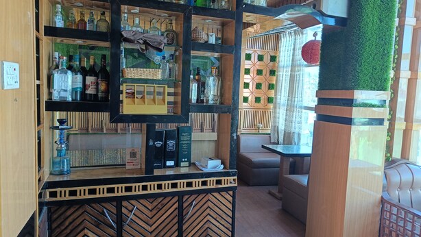 Uma área de bar com prateleiras de garrafas de bebidas, um narguilé e um espaço de estar com um banco acolchoado e uma pequena mesa.