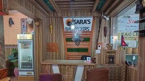 Uma área de recepção com balcão de madeira, duas poltronas de couro, um refrigerador com bebidas e um painel com a placa 'Sara's Backpackers'.
