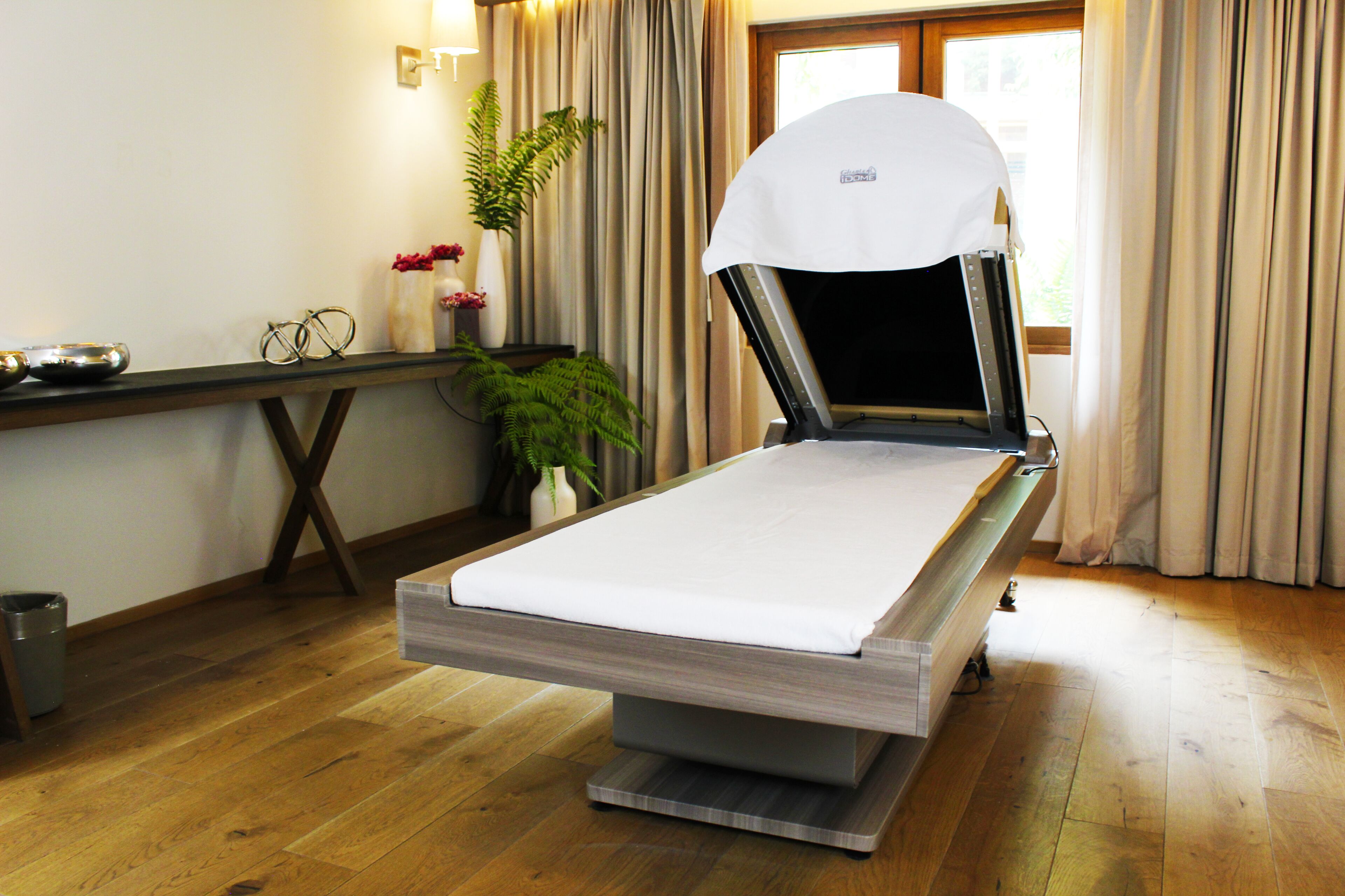 Um quarto com mesa de massagem, piso de madeira, janela com cortinas, uma planta e uma pequena mesa com vaso e copos.