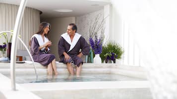 Duas pessoas sentadas em um ambiente de spa com design moderno, incluindo um banco de pedra e plantas decorativas.