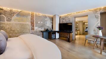 Quarto de hotel moderno com parede de destaque em pedra, TV de tela plana, pequena geladeira e área de estar aconchegante com sofá e poltrona.