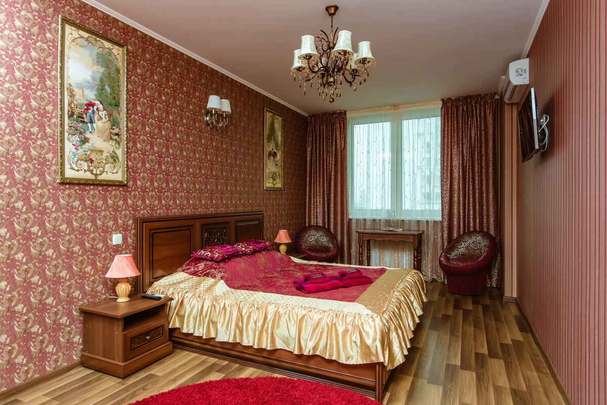 Um quarto com uma cama grande, um lustre, uma mesa de cabeceira, uma cadeira vermelha e uma janela com cortinas.