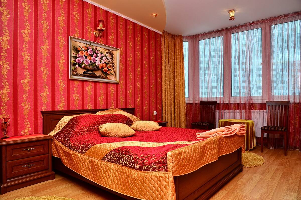 Quarto com cama grande, mesa de cabeceira, mesa auxiliar, quadro emoldurado e janela com cortinas.