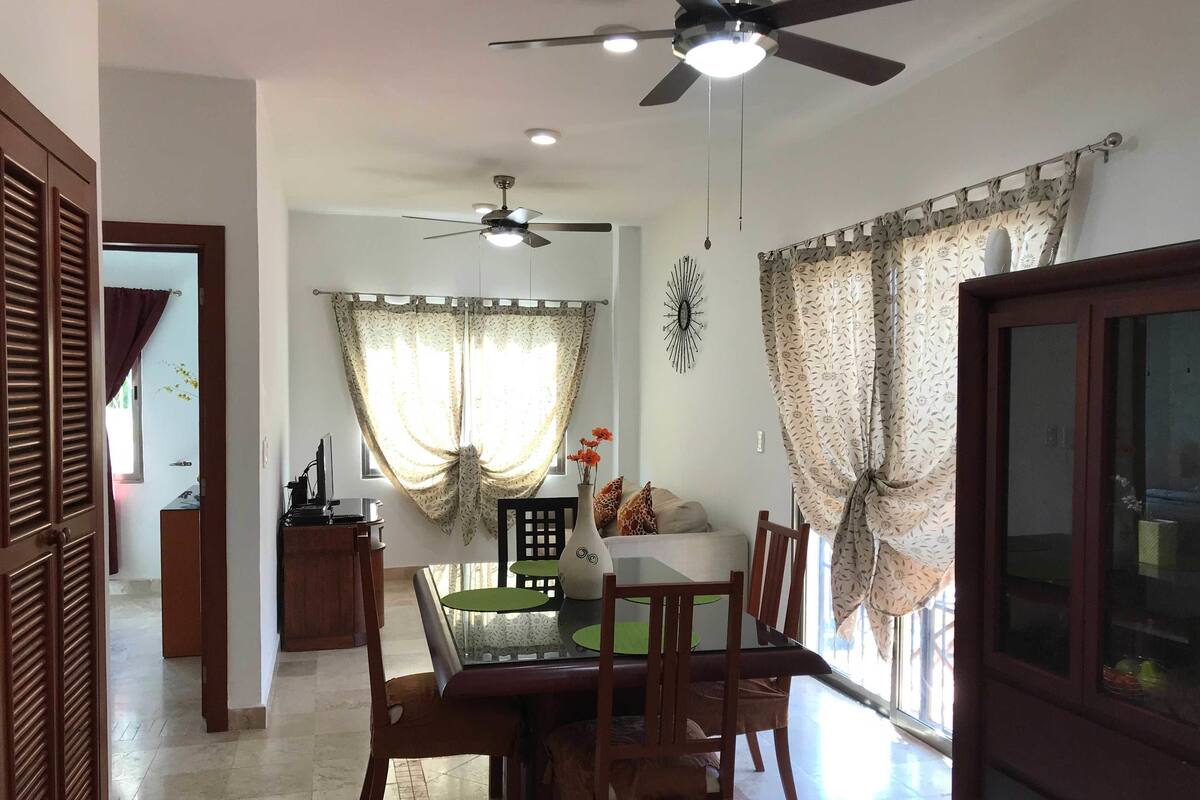 Uma área de refeições com mesa, cadeiras, ventilador de teto e cortinas.