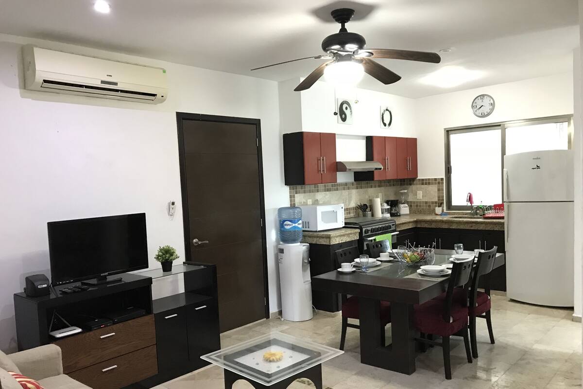 Cozinha moderna com micro-ondas, fogão e frigorífico. Há uma mesa de jantar com cadeiras e uma pequena mesa com um objeto decorativo. Um ventilador de teto é visível acima da cozinha.