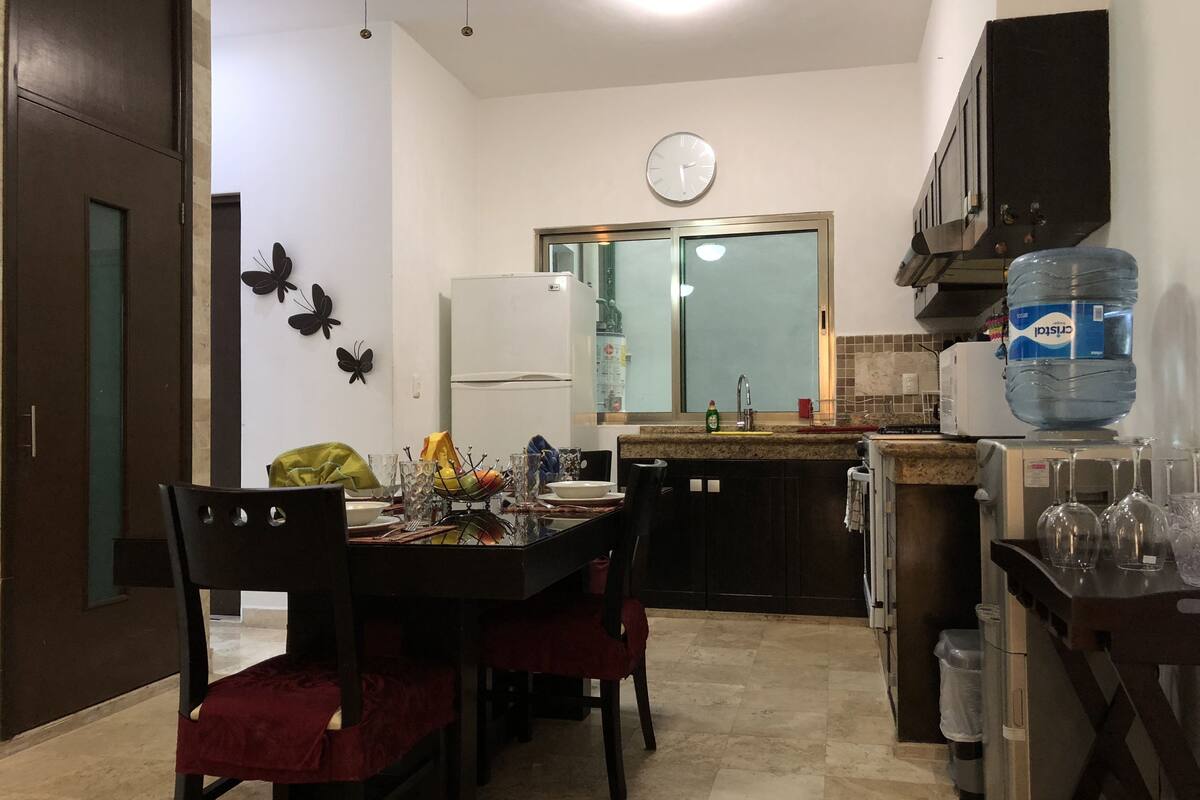 Cozinha compacta com área de refeições, geladeira, dispenser de água e um relógio na parede.