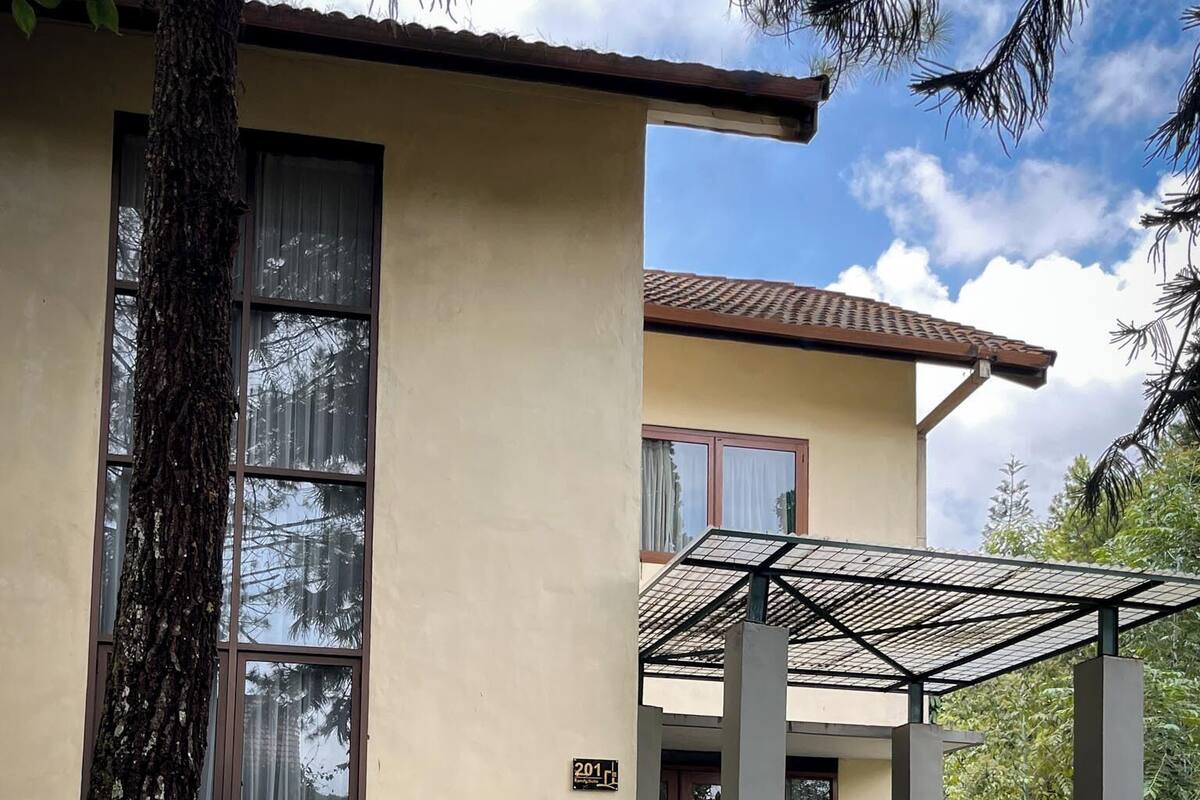 Edifício de dois andares com base em pedra e seção superior bege. Possui uma grande janela no primeiro andar e uma janela menor no segundo. Uma escadaria de pedra leva à entrada, e uma área coberta com telhado de metal é visível.