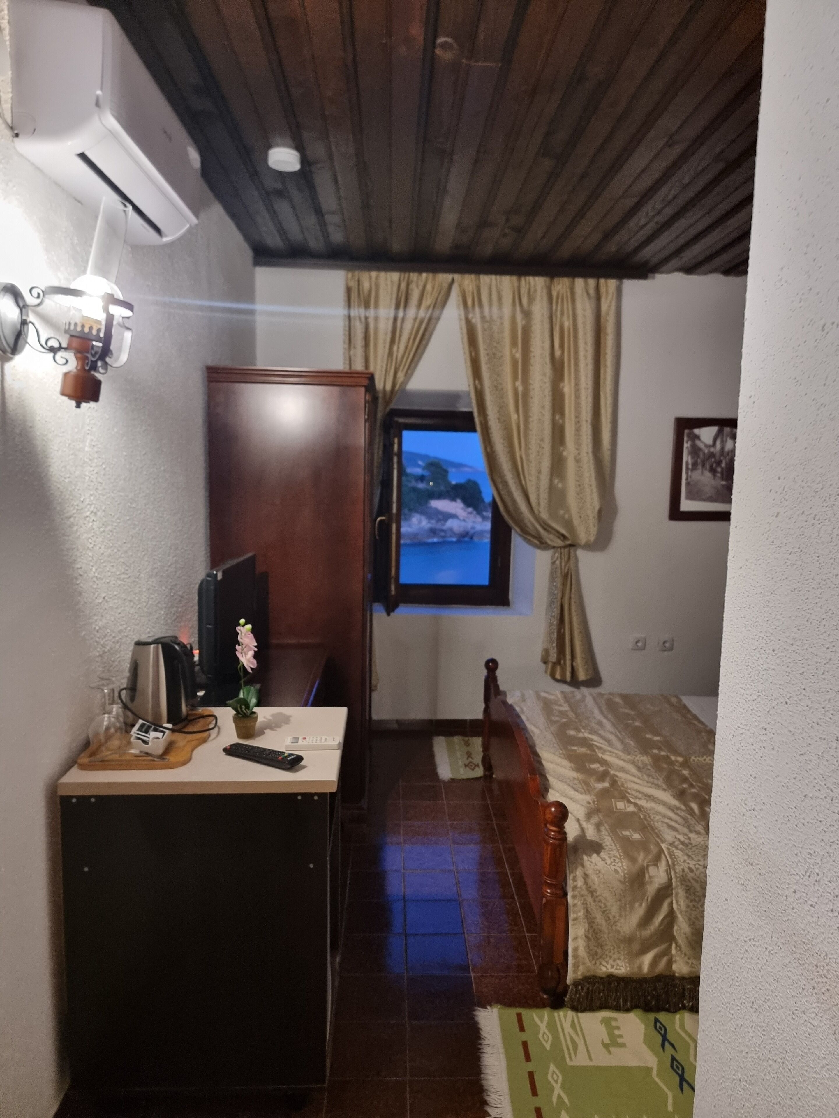 Quarto pequeno com uma cama, uma escrivaninha com um computador, uma janela com cortinas e um ar condicionado fixado na parede.