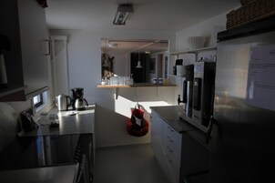 Uma cozinha compacta com cafeteira, micro-ondas e geladeira. Há uma janela que oferece iluminação natural, e uma pequena prateleira com copos.