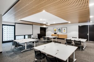 Uma sala de conferências moderna, com mesas e cadeiras brancas, uma tela de projetor e teto de madeira.