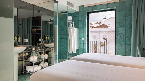 Quarto de hotel moderno com uma cama grande, banheiro com paredes de vidro e varanda com vista para edifícios.