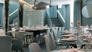 Uma sala de jantar moderna com mesas e cadeiras de vidro, uma escada ao fundo e um espelho grande na parede.
