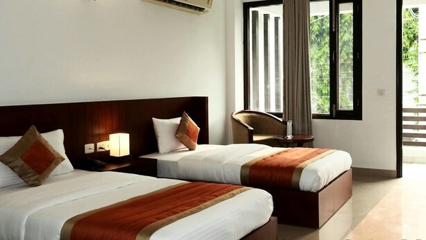 Quarto de hotel com duas camas, um ventilador de teto, um ar condicionado, uma mesa de cabeceira com abajur, uma cadeira e vista para o verde lá fora.
