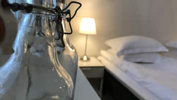 Uma garrafa de vidro transparente com fecho metálico, uma mesa de cabeceira branca, um abajur e uma cama com roupa de cama branca em um quarto de hotel.