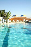 Piscina com água cristalina, rodeada por espreguiçadeiras e guarda-sóis, e um edifício com telhado cerâmico ao fundo.