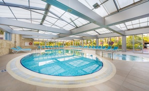 Uma grande piscina interior com várias pistas de natação, rodeada por espreguiçadeiras azuis e um teto de vidro que permite a entrada de luz natural.