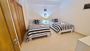 Quarto com duas camas, guarda-roupa de madeira, mesa de cabeceira com abajur e uma pequena mesa.