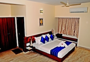 Quarto de hotel com cama, mesa de cabeceira, escrivaninha, janela com cortinas, ventilador de teto e ar condicionado.