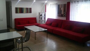 Uma sala de estar com um sofá vermelho, uma mesa de centro branca e uma cadeira preta. Há uma estante com diversos objetos e um quadro na parede.