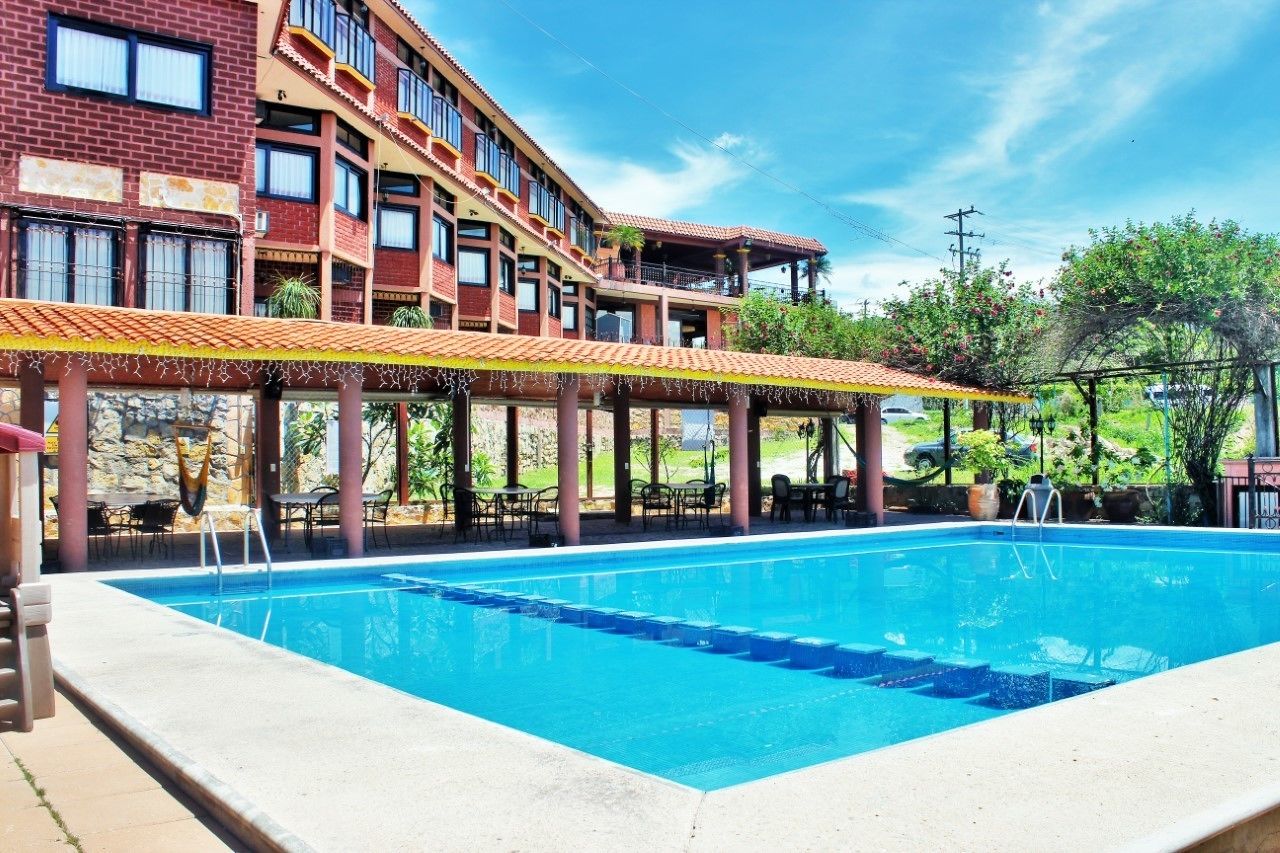Piscina com área coberta, espaço para sentar ao ar livre e um edifício com vários andares ao fundo.