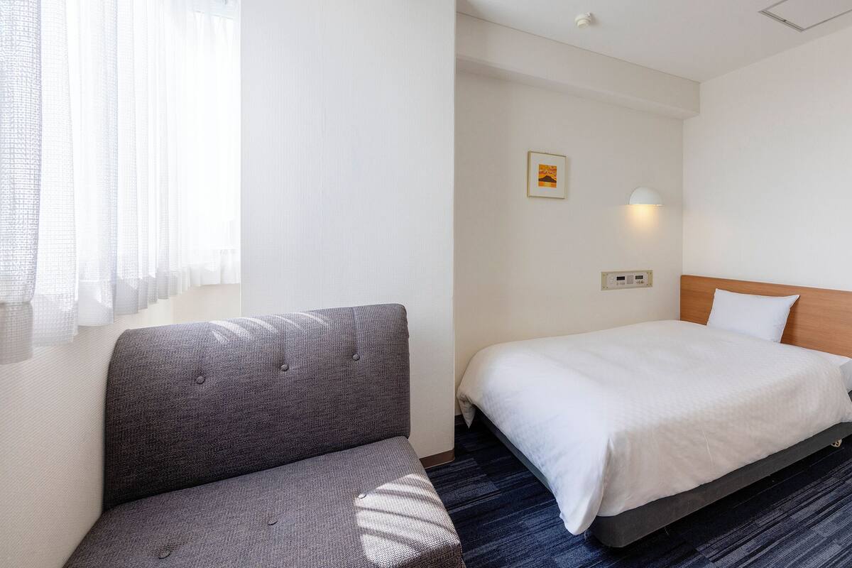 Quarto de hotel com uma cama, um sofá, uma luminária fixada na parede e uma pequena imagem.