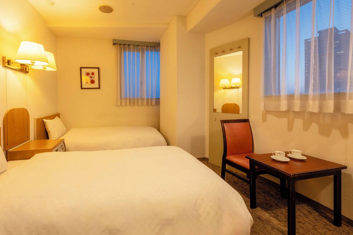 Quarto de hotel com duas camas, uma mesinha com chávenas, uma cadeira, uma janela com cortinas e um espelho fixado na parede.