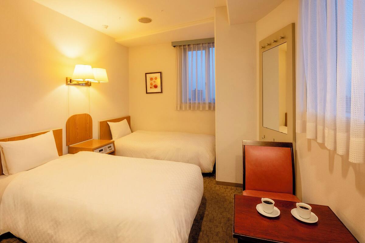 Quarto de hotel com duas camas, uma mesa de cabeceira com abajur, um espelho e uma janela com cortinas.