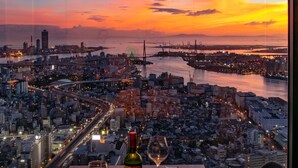 Uma mesa com uma garrafa de vinho e copos, com vista para um horizonte urbano com o pôr do sol sobre a água.