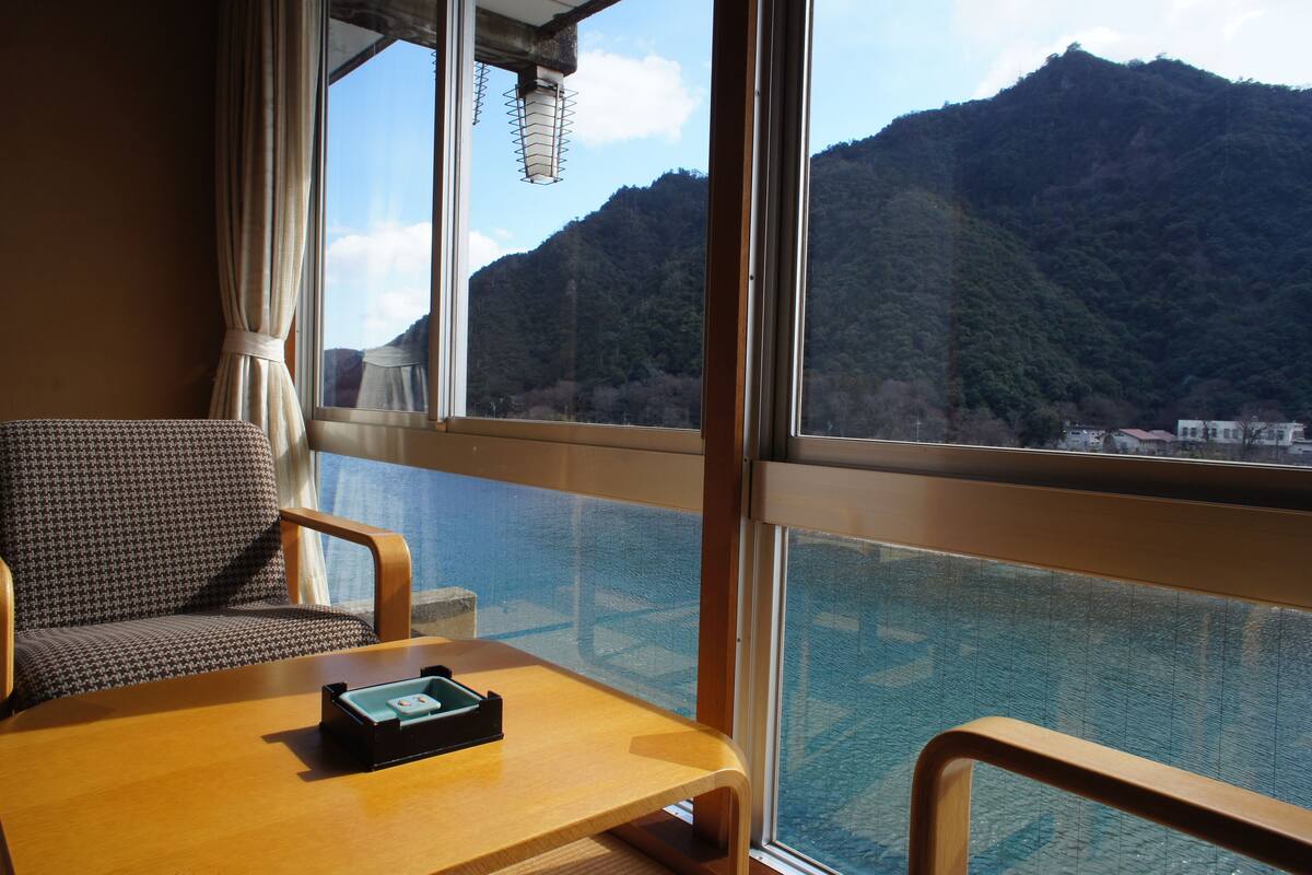 Quarto com mesa de madeira, cadeira estofada e uma grande janela com vista para uma paisagem montanhosa.