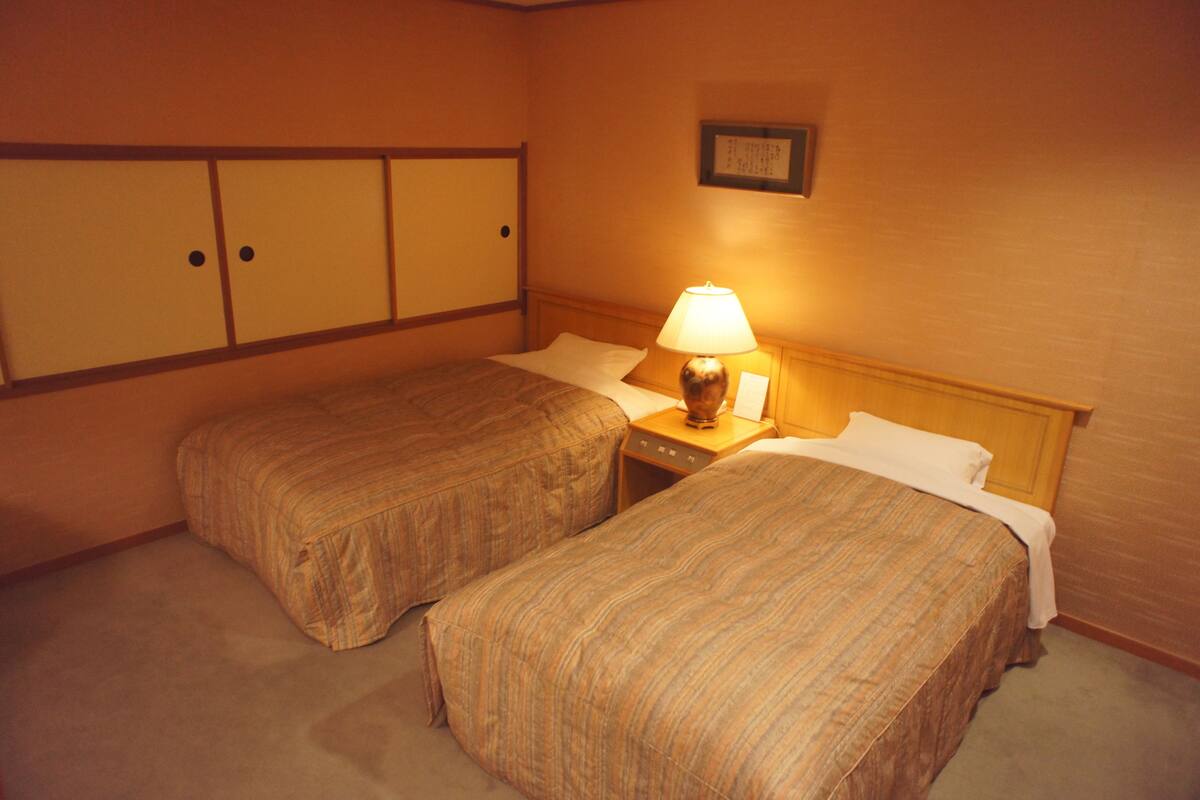 Quarto de hotel com duas camas de solteiro, uma mesa de cabeceira com abajur e cabeceira de madeira.