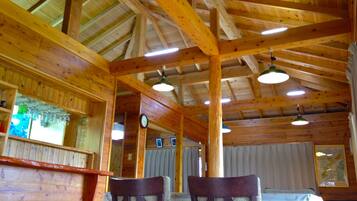 Interior em madeira com teto alto, vigas aparentes e luminárias pendentes. Há duas cadeiras e uma mesa com toalha branca.