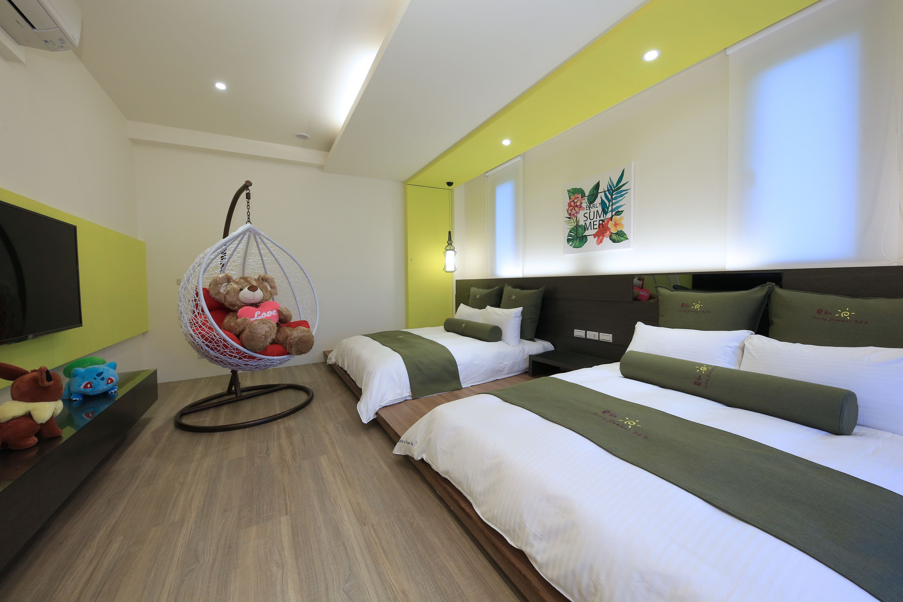 Quarto de hotel com duas camas, uma cadeira suspensa com brinquedos de pelúcia, uma televisão de tela plana e uma parede com arte tropical.