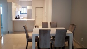 Uma área de refeições moderna com mesa branca e cadeiras cinzas, cozinha ao fundo e porta que dá para outro quarto.