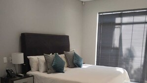 Quarto moderno com uma cama grande, mesa de cabeceira com abajur e janela com persianas.