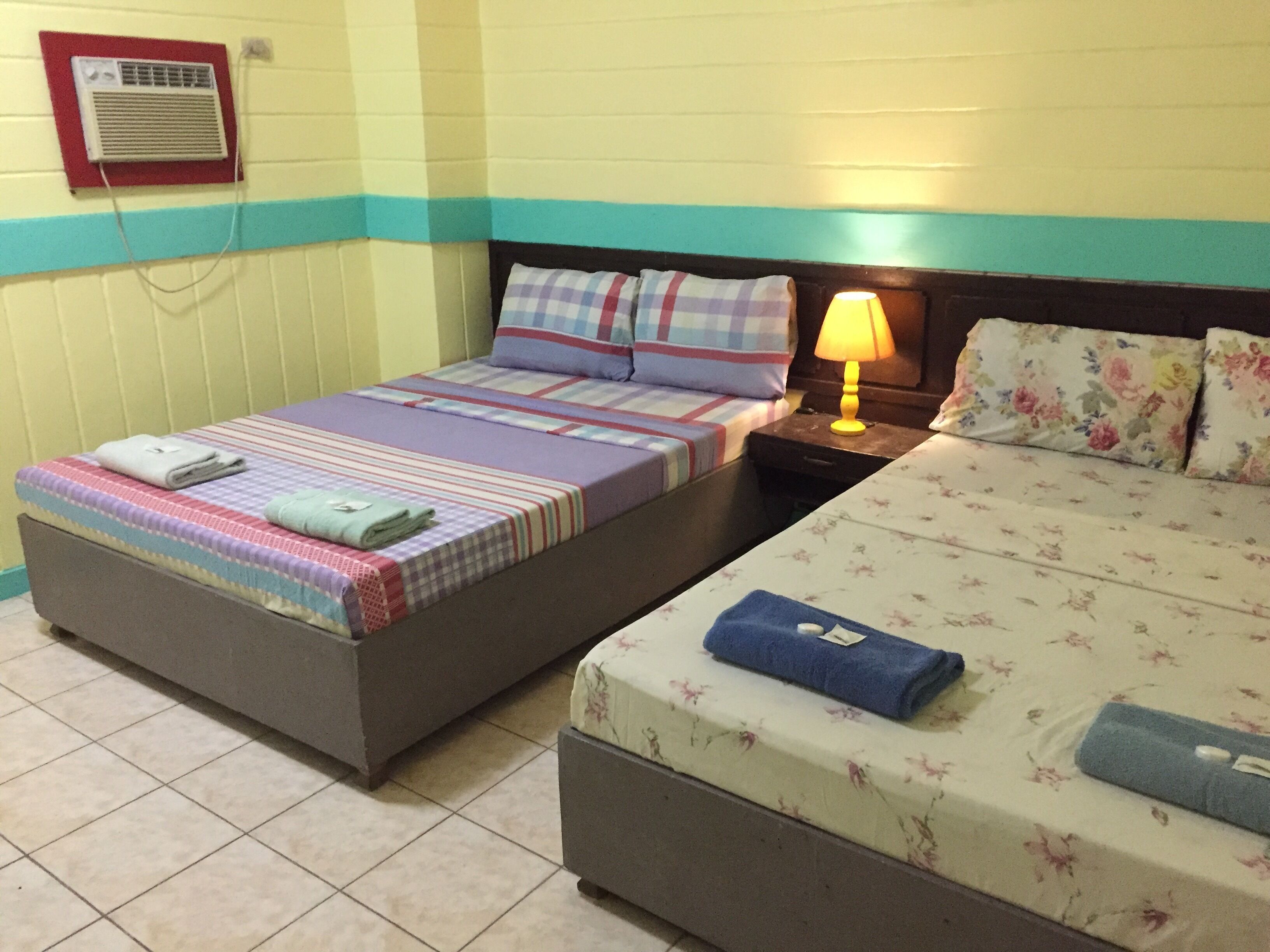 Quarto com duas camas, uma com colcha florida e outra com colcha listrada. Há uma mesa de cabeceira com uma luminária entre as camas.