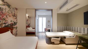 Quarto de hotel com cama, sofá-cama, uma mesinha e televisão. Há uma porta que dá para outro quarto e uma janela com persianas.