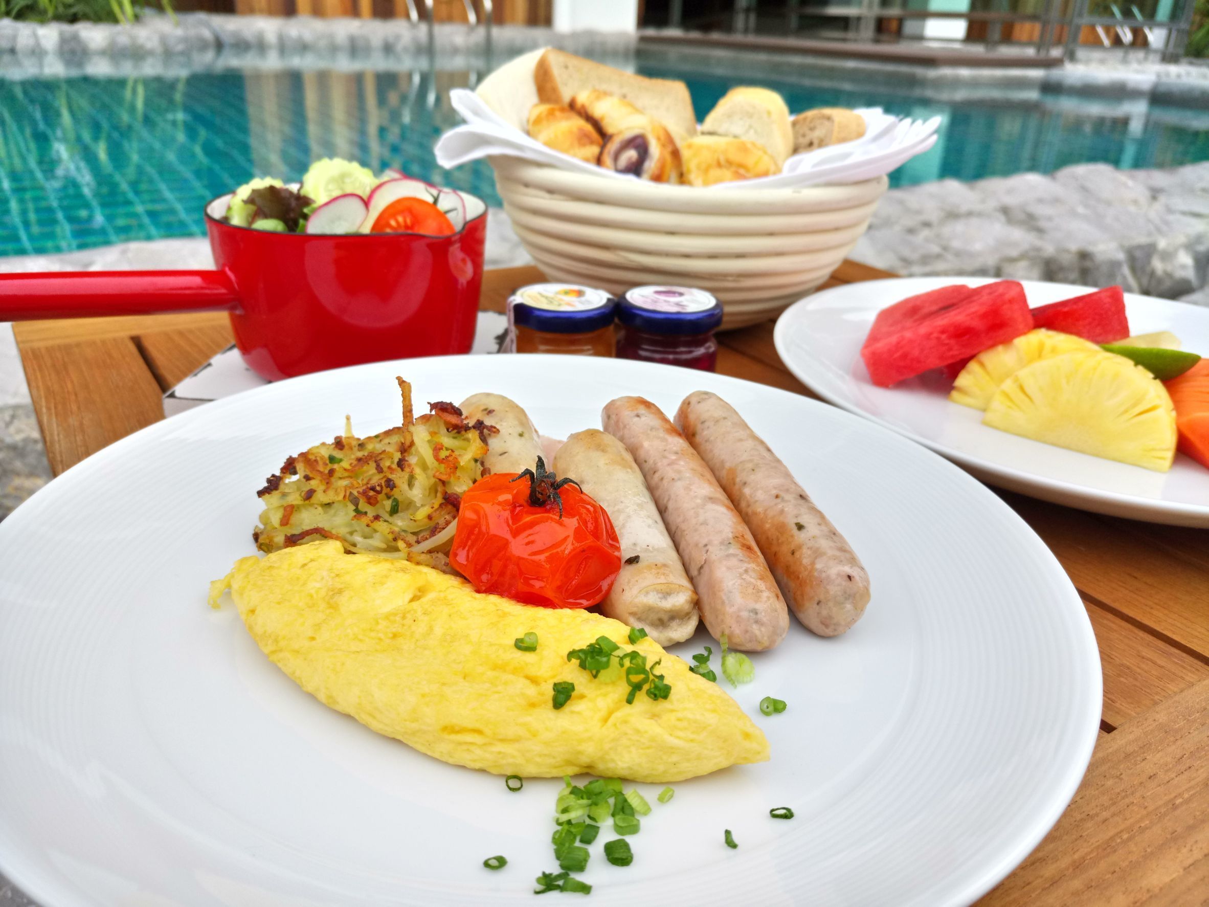 Prato de café da manhã com ovos, linguiça e legumes, uma tigela de pão e um prato de frutas à beira da piscina.
