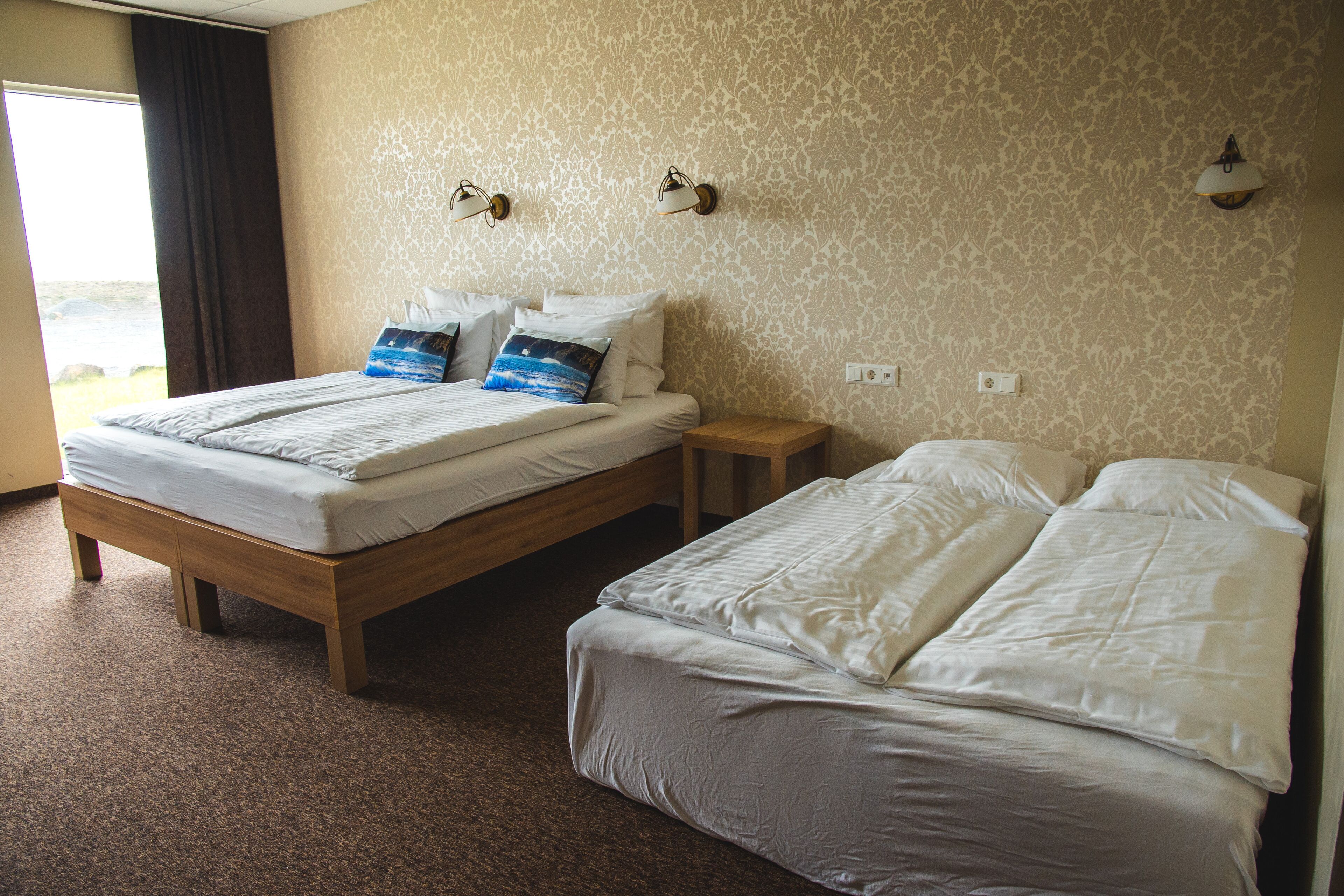Quarto de hotel com duas camas, uma mesinha de cabeceira de madeira e uma janela com cortinas.