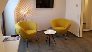 Quarto moderno com duas poltronas amarelas, uma pequena mesa branca e um abajur. Uma TV de tela plana está fixada na parede.
