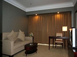 Quarto de hotel com sofá bege, mesa de centro redonda pequena, mesa lateral de madeira com abajur e televisão em um móvel.