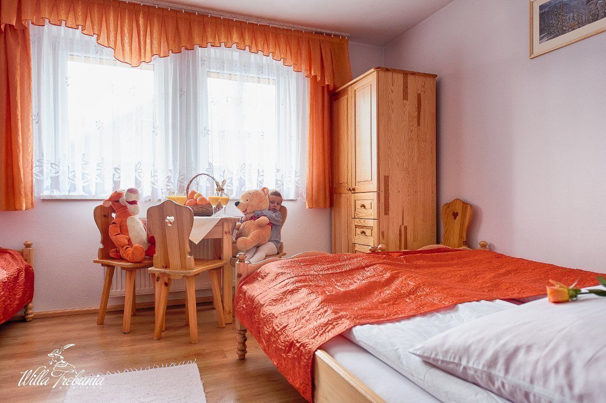 Quarto com cama, guarda-roupa de madeira, escrivaninha com cadeiras e janela com cortinas.