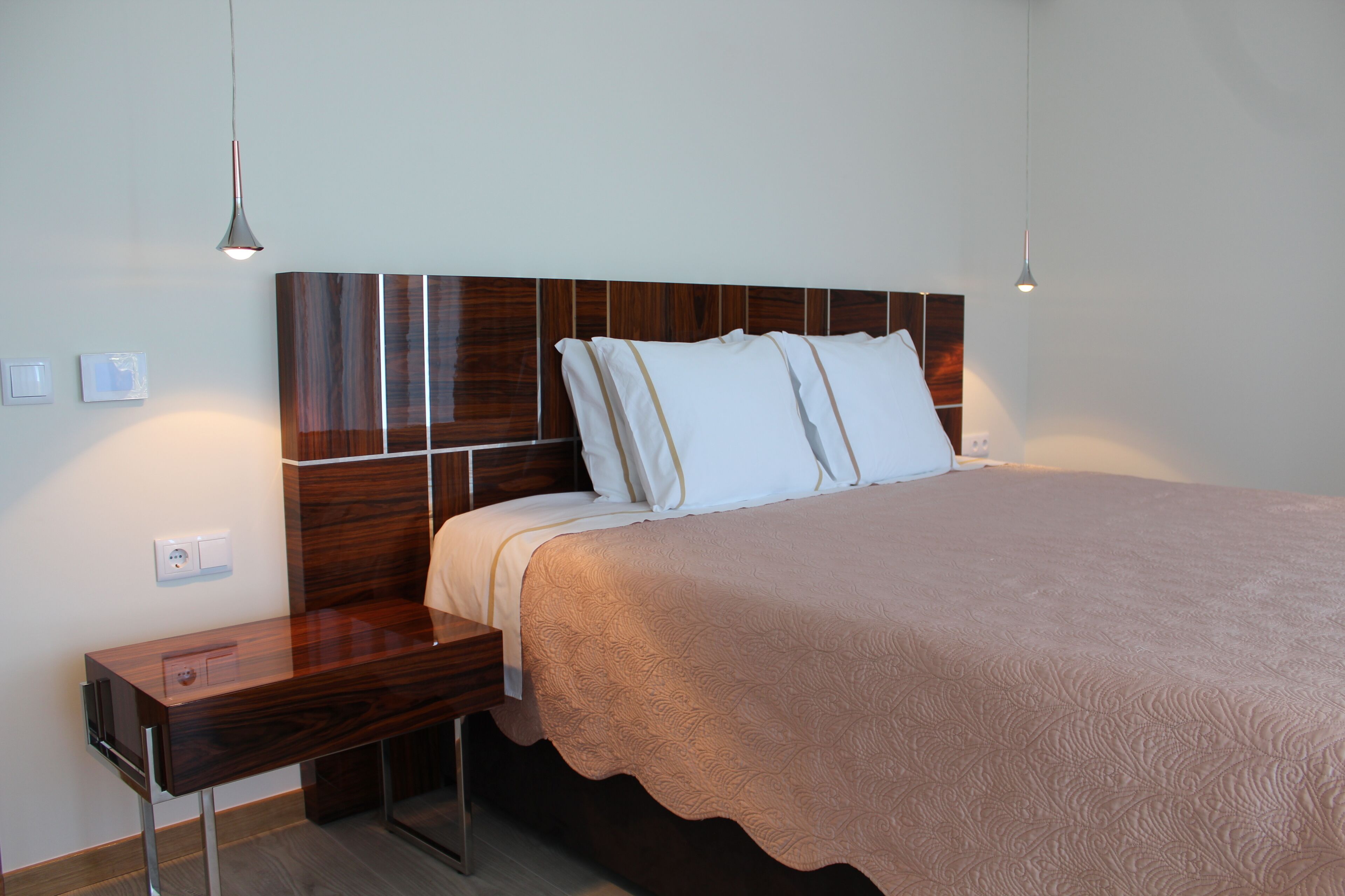 Quarto moderno com cabeceira de madeira, mesa de cabeceira e cama com colcha bege.